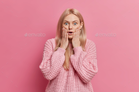 Stunned blonde woman stares with shock at camera keeps hands on cheeks ...