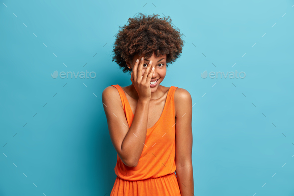 Happy shy woman makes face palm looks through fingers dressed in orange ...