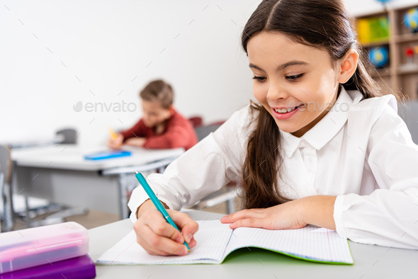 Smiling schoolgirl writing in notebook during lesson in classroom Stock ...