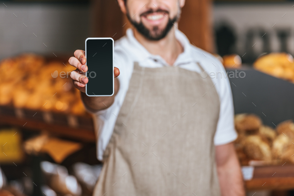 partial view of smiling shop assistant showing smartphone with blank ...