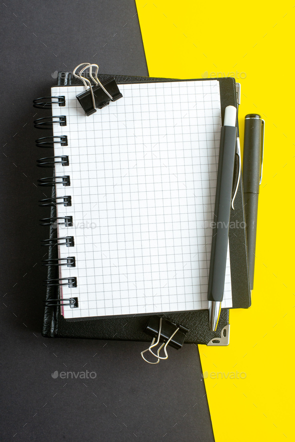 Vertical view of spiral notebook on book and pens on black yellow ...