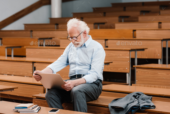 grey hair professor sitting on desk in empty lecture room and using ...