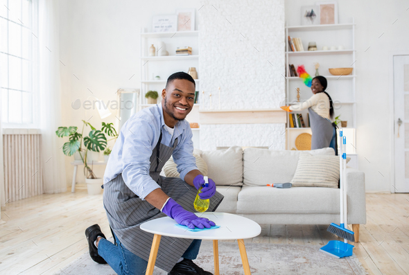 Smiling black guy wiping table in living room while his girlfriend ...