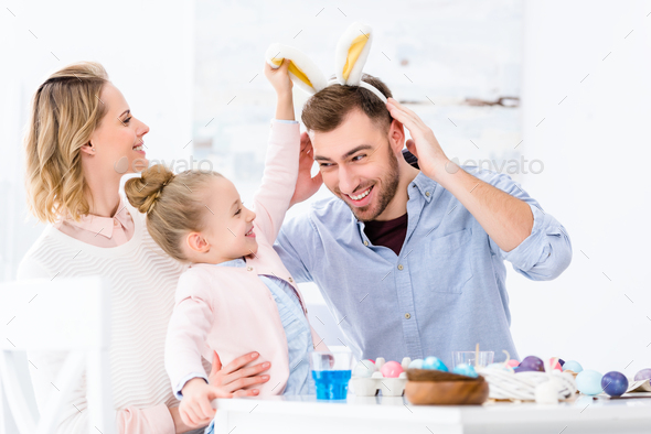 Smiling family having fun with bunny ears by table with painted eggs on ...