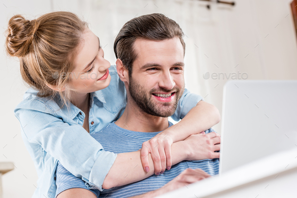 Smiling young couple hugging while using laptop at home Stock Photo by ...