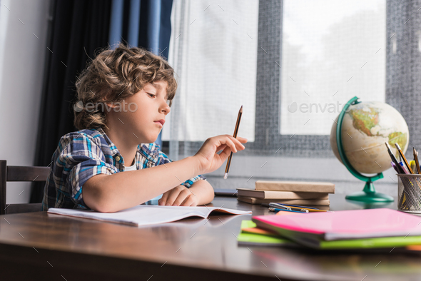side view of thoughtful little boy looking away while doing homework at ...