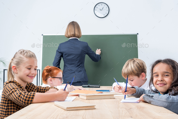 back view of teacher writing on chalkboard while students studying in ...