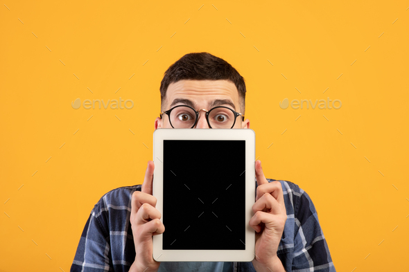 Shocked millennial guy hiding behind tablet computer with empty screen ...