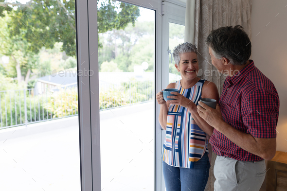Happy senior caucasian couple standing by window talking holding cups ...