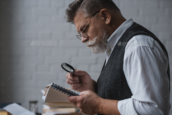 handsome senior man reading notes in notebook with magnifying glass ...