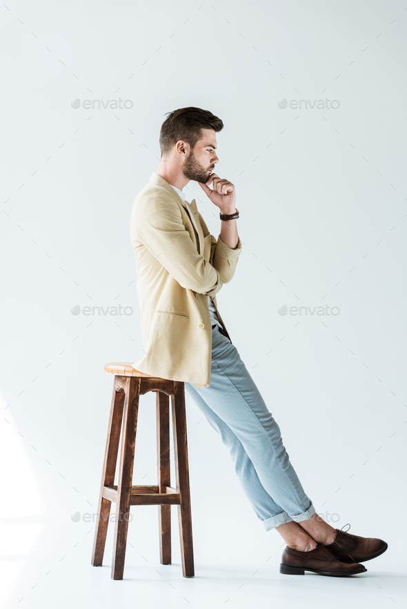 Stylish young man thinking while sitting on stool on white background ...