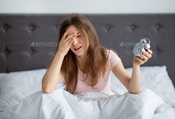 Depressed young woman with alarm clock sitting on bed, crying in ...