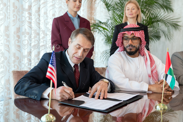 A group of people sitting by table while one of them signing contract ...