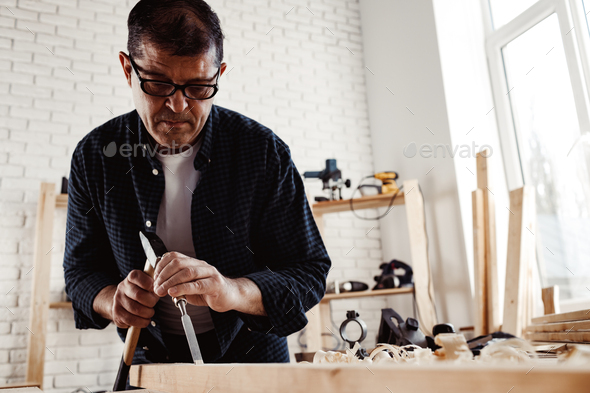 Middle-aged man carpenter working in a workshop with chisel and hammer ...