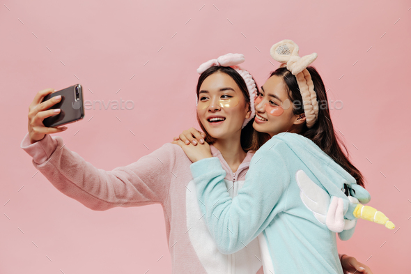 Tanned cool young women in pajamas take selfie on pink background ...