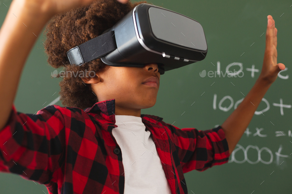 African american schoolboy in front of chalkboard in classroom using vr ...