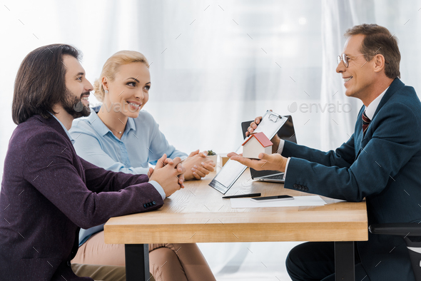 adult smiling insurance agent showing contract and house model to young ...