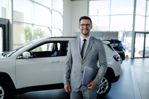 Confident young car dealer standing in showroom and holding laptop ...