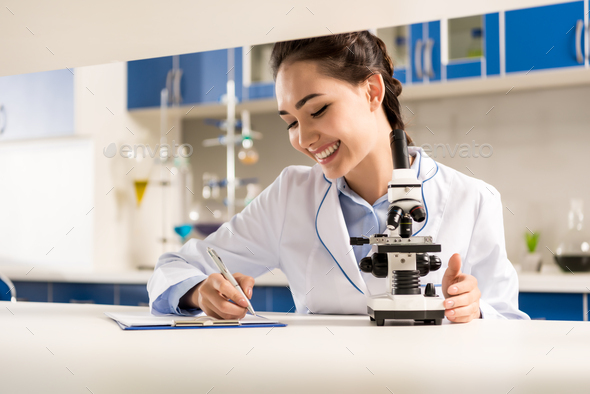 Young smiling lab technician taking notes after doing microscope sample ...