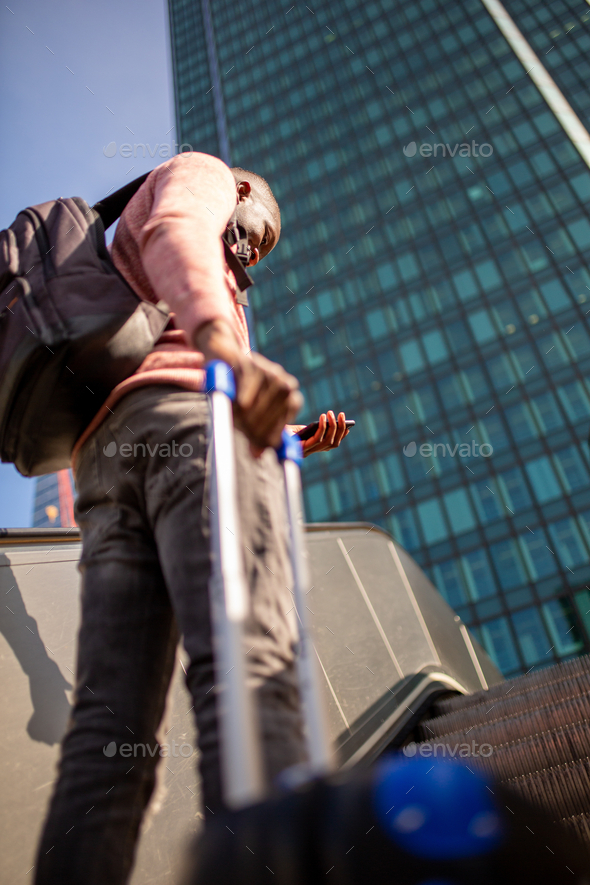 behind young black man going up escalator with suitcase and mobile ...