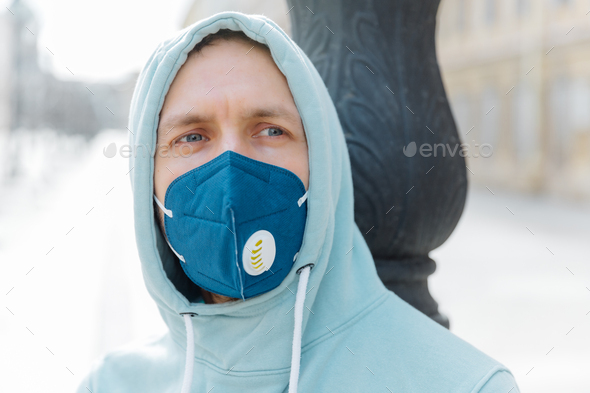 Headshot of thoughtful young man wears hood and respiratory mask, walks ...