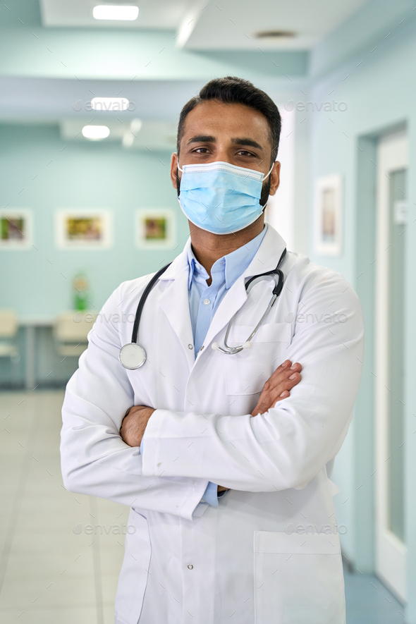 Male happy indian doctor wearing facemask standing in clinic hospital ...