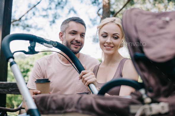 smiling parents sitting on bench near baby carriage in park and holding ...