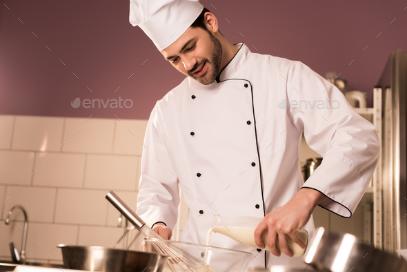 portrait of confectioner in chef hat pouring milk into bowl while ...
