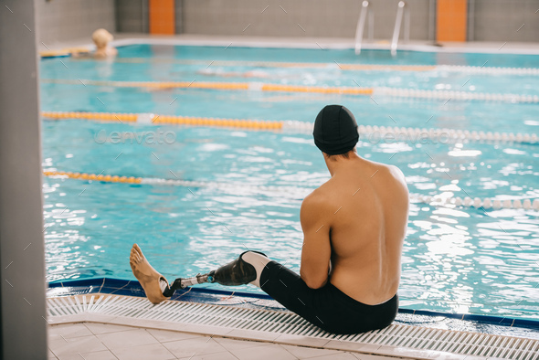 rear view of young swimmer with artificial leg sitting on poolside of ...