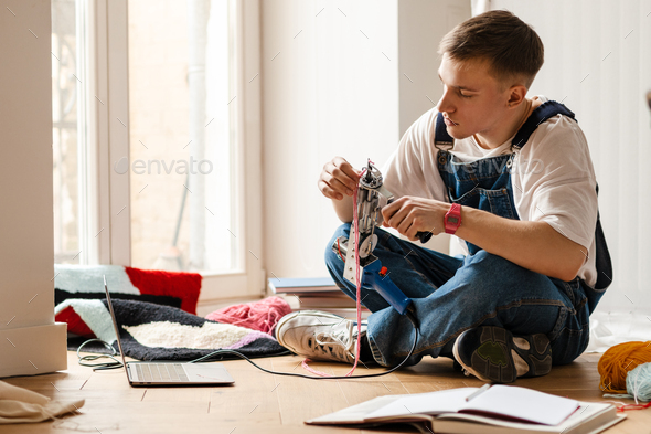 Young man using laptop and sewing machine while working on craft rug ...