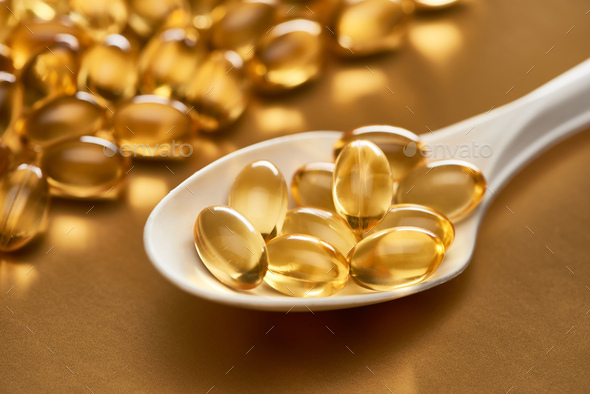 close up view of shiny fish oil capsules in spoon on golden background ...