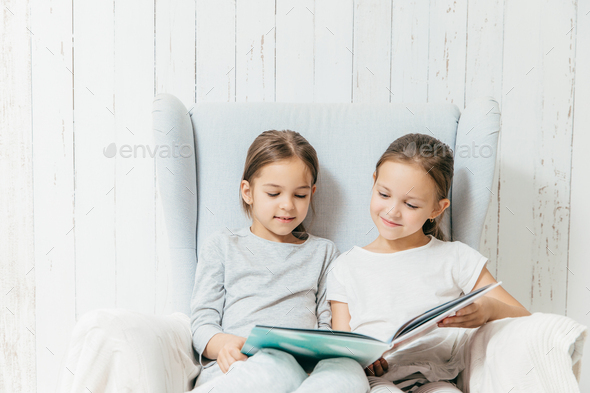 Two adorable sisters sit in sofa, read interesting book, sit in ...