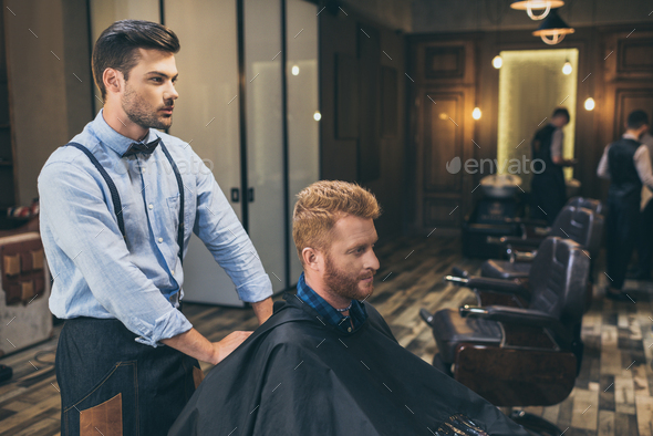 Stylish young barber standing behind customer that sitting in barber ...
