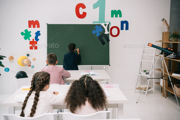 Back view of pupils sitting at desks and looking at classmate writing ...