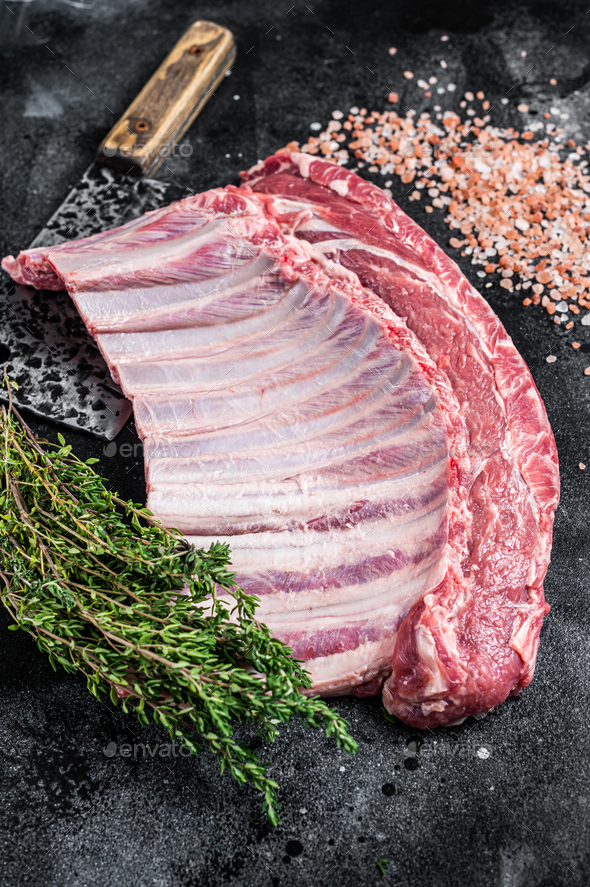 Raw rack of lamb ribs on butcher cutting board with herbs. Black ...