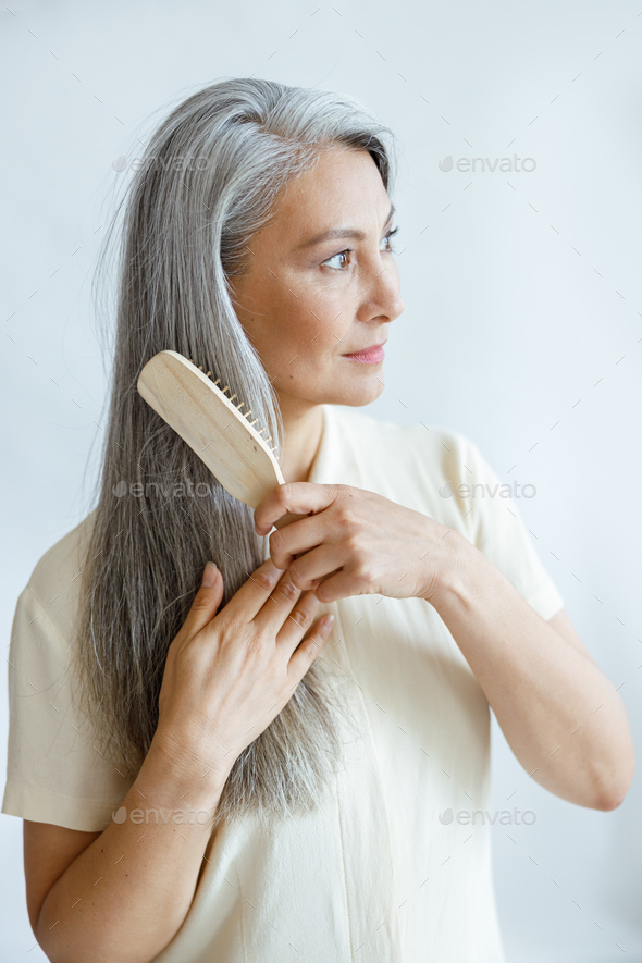 Languid middle aged Asian woman brushes natural hoary hair on light ...