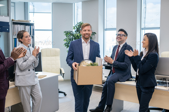 Cheerful employee carrying box with supplies among colleagues clapping ...