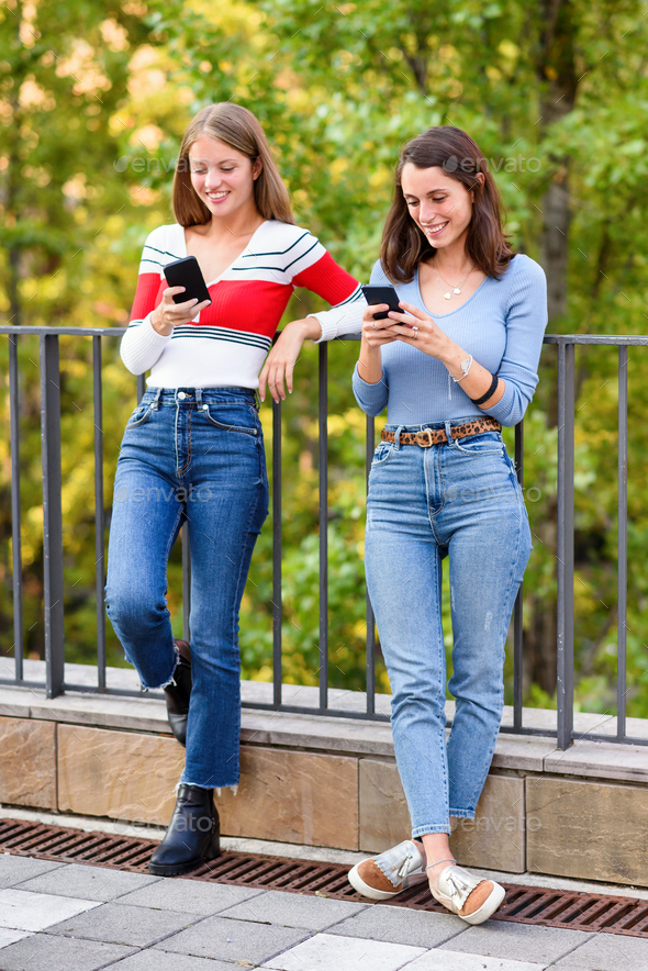 Two happy young woman standing using their mobile phone Stock Photo by ...