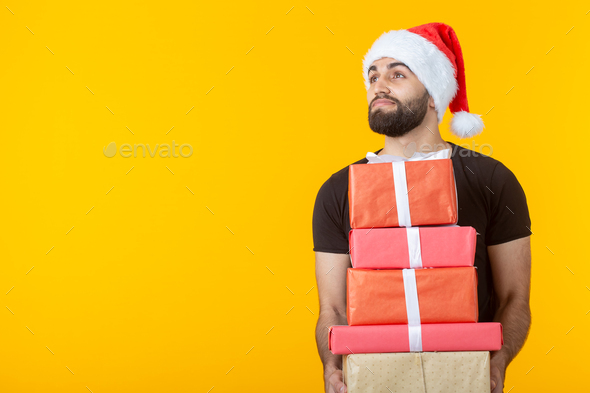 Disgruntled young man with a beard in a Santa Claus hat holds five gift ...