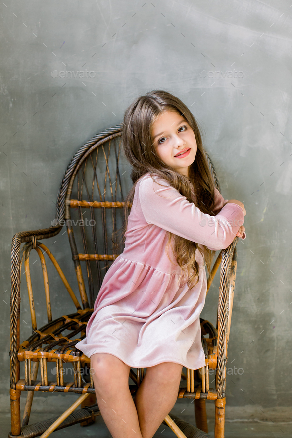 Pretty small girl in beautiful pink dress sits on the wooden chair and ...