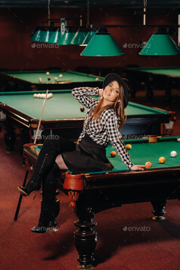 A girl in a hat in a billiard club sits on a billiard table with a cue ...