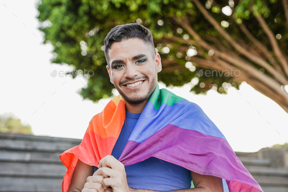 Young transgender man smiling on camera holding rainbow flag outdoor ...