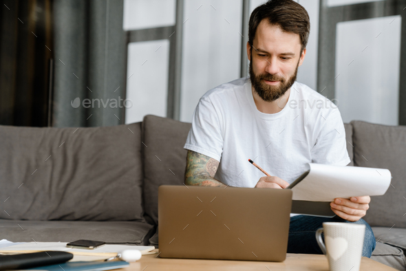 Bearded european man writing down notes while working with laptop Stock ...