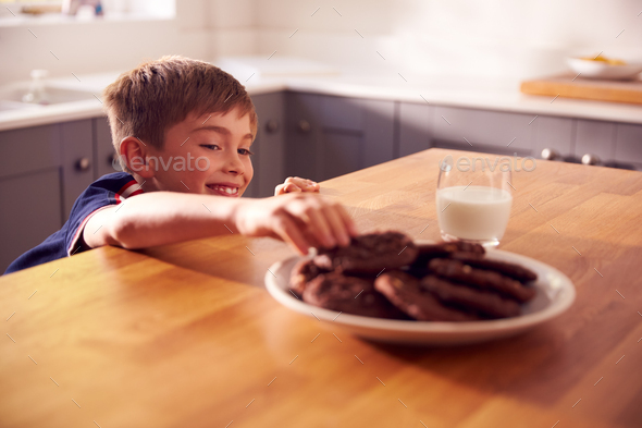 Boy At Home In Kitchen Reaching Up To Take Cookie From Plate On Counter ...