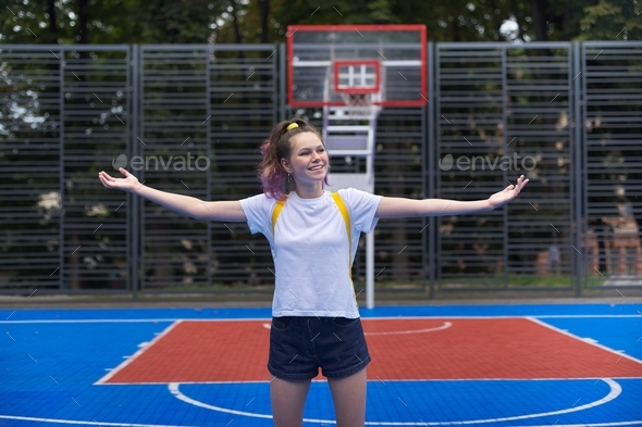 Active smiling teenage girl on street basketball court with arms raised ...