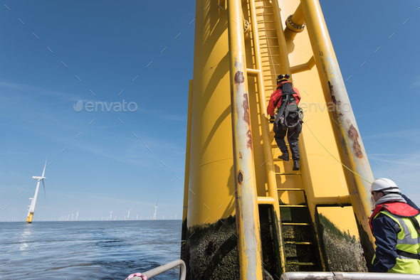 Engineers climbing wind turbine ladder from boat at offshore windfarm ...