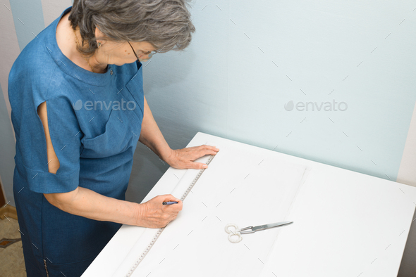 Elderly woman is sewing indoors. Senior dressmaker designing on fabric ...