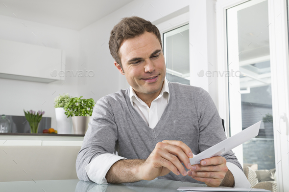 Smiling man sitting in kitchen opening envelope Stock Photo by westend61