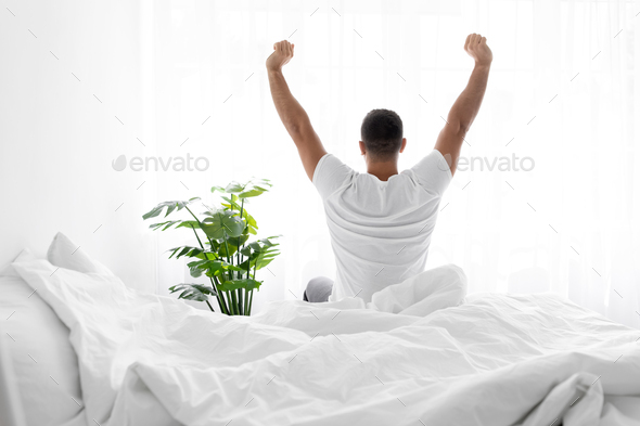 Happy young caucasian muscular man sits on white bed, wake up ...
