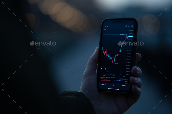 Trader holding smartphone with financial chart and analyzing price flow ...